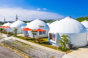 Fileira de edifícios em forma de cúpula, com mesas de piquenique e guarda-sóis vermelhos, cercada por vegetação.