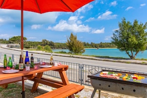 Montagem para piquenique com churrasqueira, mesa e garrafas, com vista para a água e uma estrada.