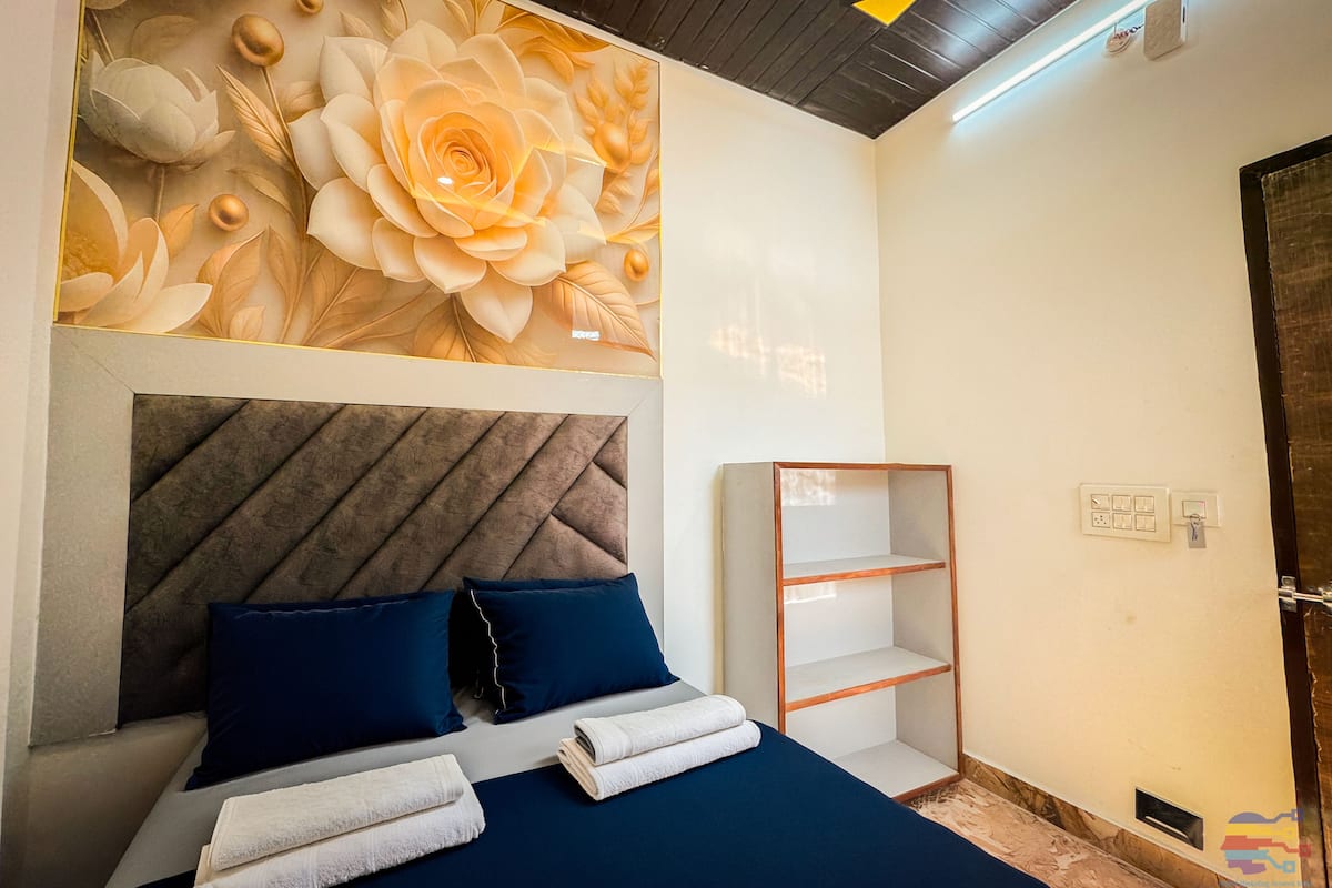 Quarto com cama, almofadas, cabeceira, parede com mural floral, prateleira e porta.