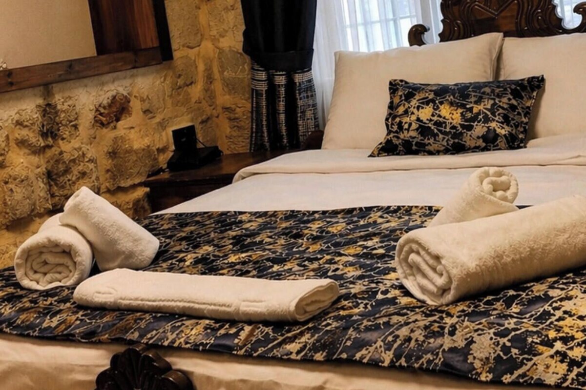 Quarto com parede de pedra, cama com colcha estampada, cabeceira de madeira e janela com cortinas leves.