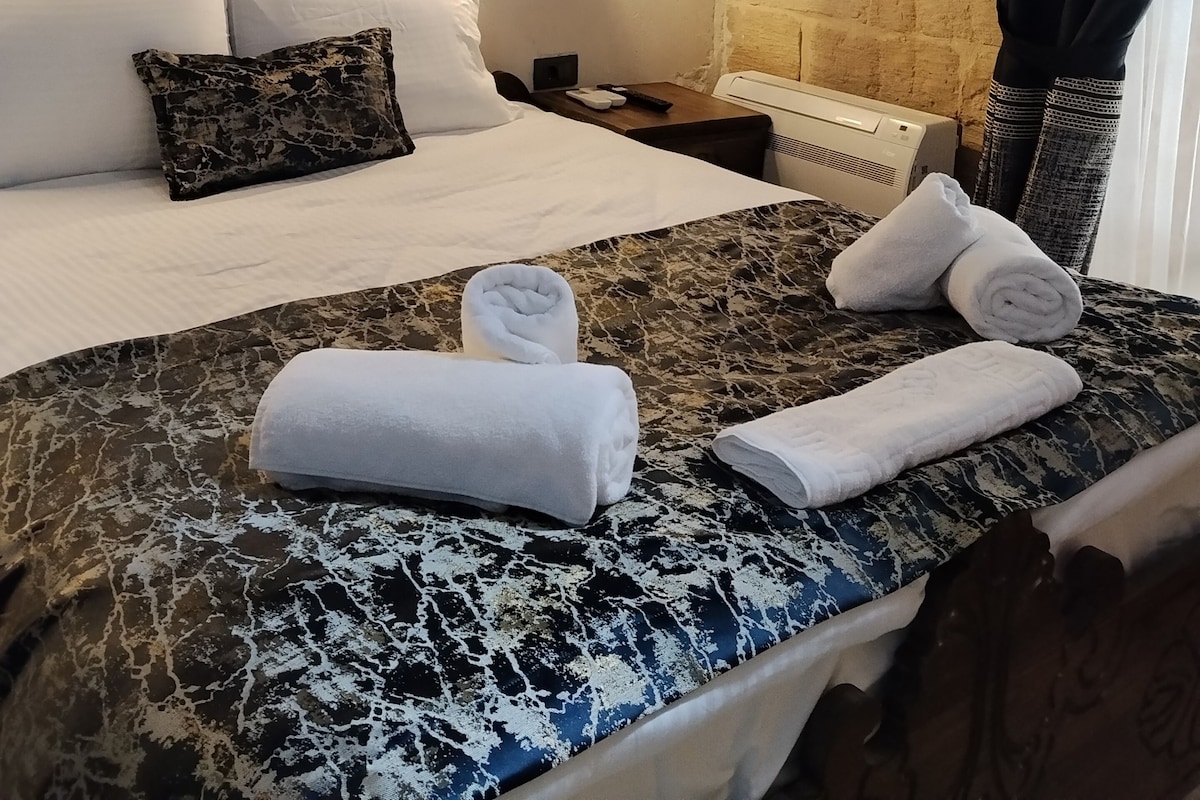 Quarto de hotel com cama, mesa de cabeceira, luminária na parede e janela com cortinas.