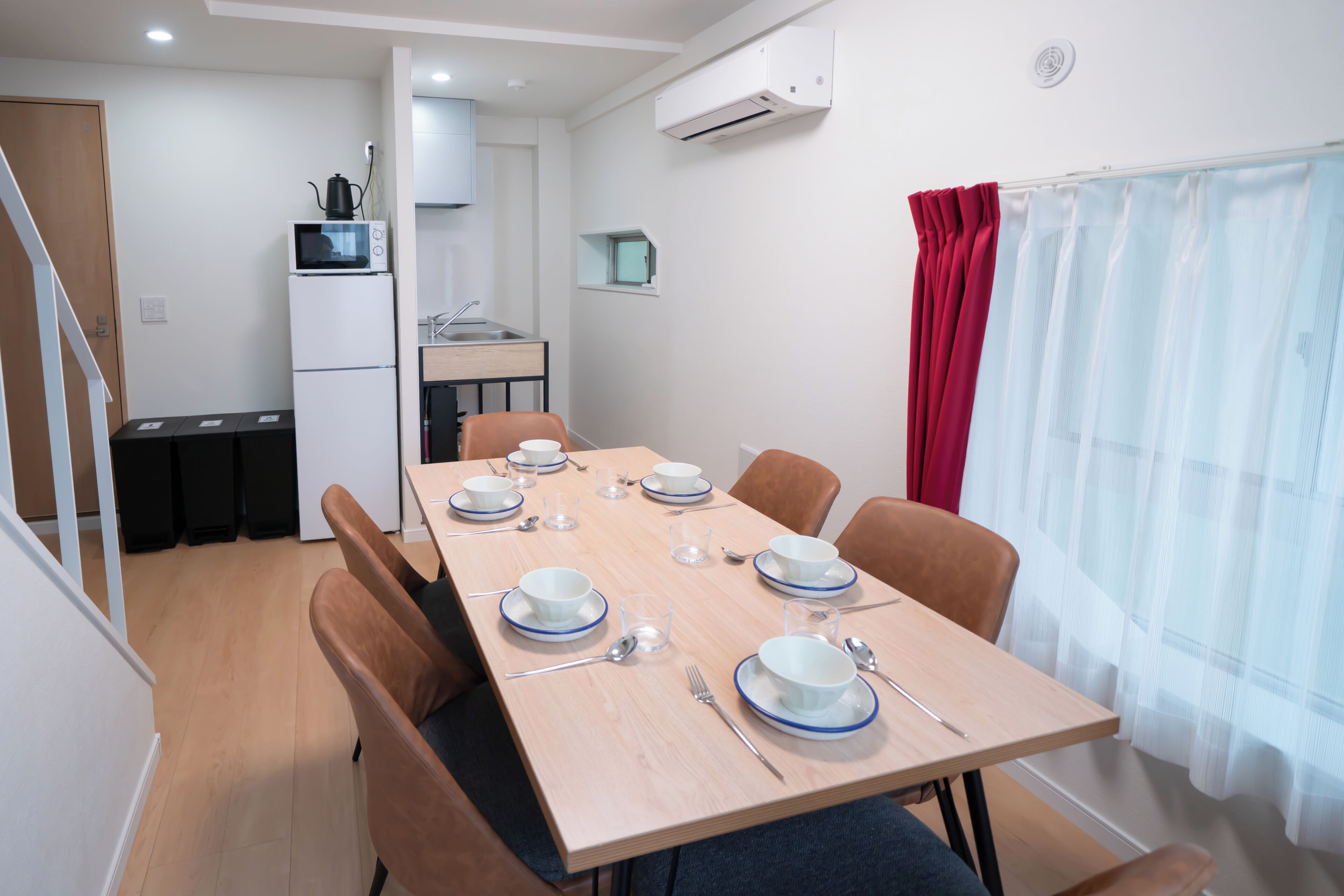 Uma sala de jantar com mesa posta para quatro pessoas, cadeiras, micro-ondas, pequena área de cozinha e janela com cortina.