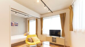 Quarto moderno com cama, poltrona amarela, mesa de centro de vidro, TV em móvel e janelas amplas com cortinas.