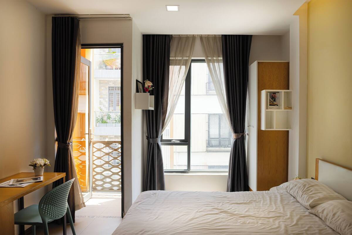 Quarto de hotel com uma cama, uma escrivaninha com cadeira, uma janela com cortinas e um armário de madeira.