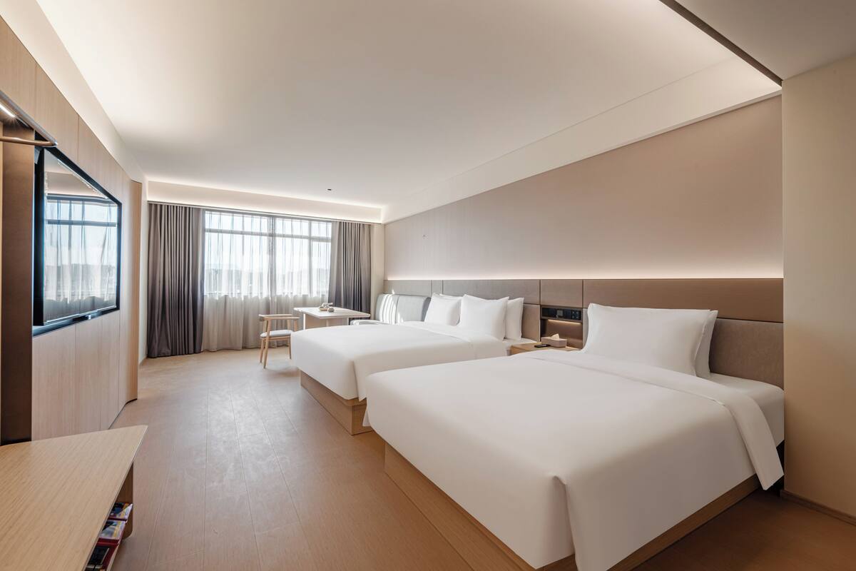 Quarto de hotel com duas camas, uma escrivaninha, uma cadeira, uma televisão e uma janela grande.