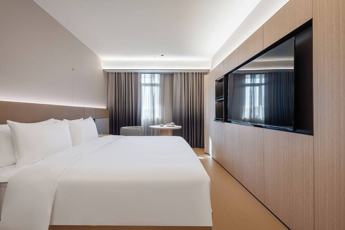 Quarto de hotel moderno com uma cama grande, uma televisão, uma mesa redonda pequena e uma cadeira.