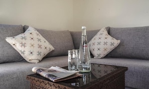 Um sofá com almofadas estampadas, uma garrafa de vidro, dois copos e uma revista sobre uma mesa de vidro.