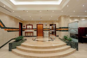 Um hall de hotel com piso de mármore, escadaria central e balcão de recepção.