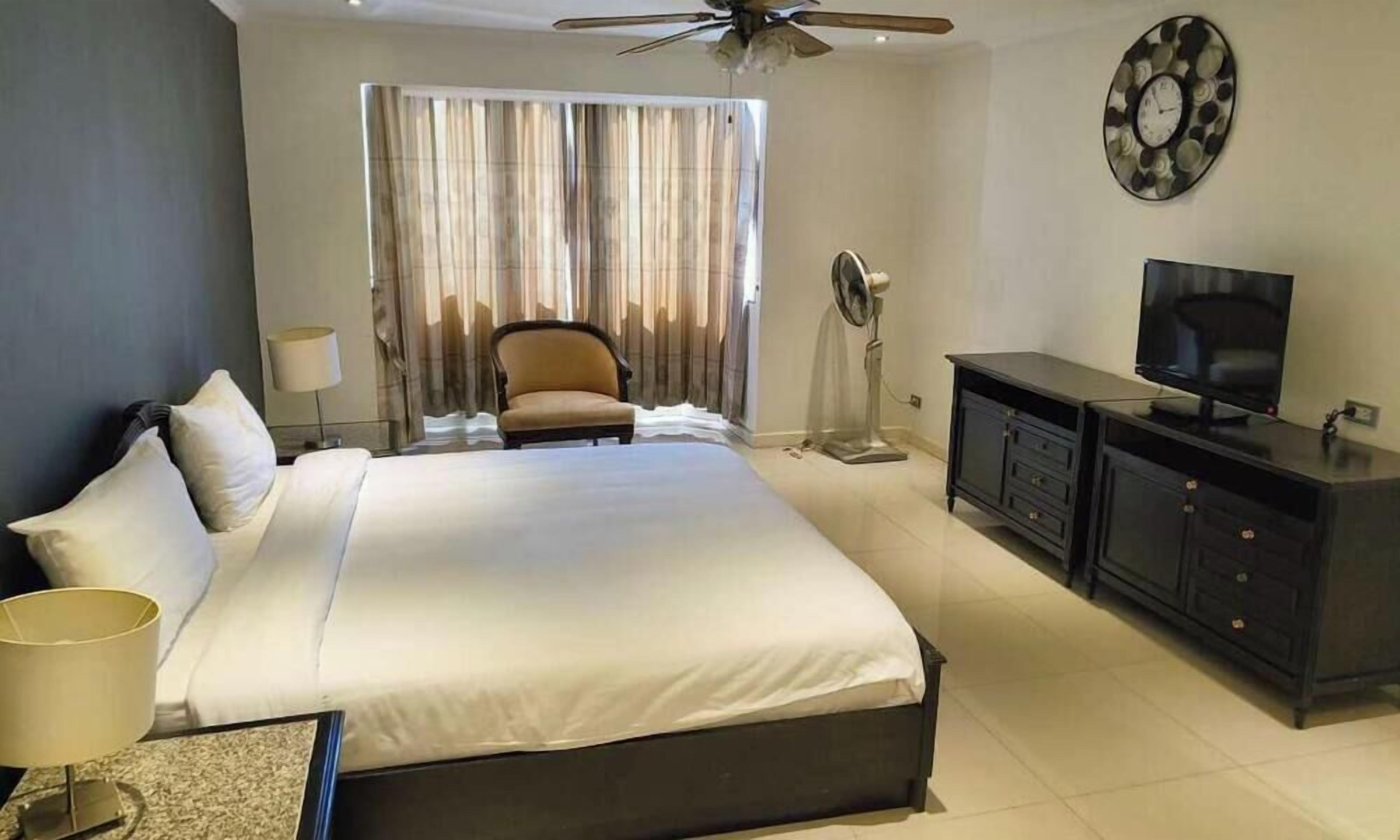 Quarto de hotel com cama, cadeira, televisão, escrivaninha e ventoinha no teto.