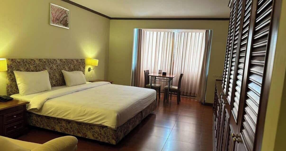 Quarto de hotel com cama, mesas de cabeceira, escrivaninha com cadeiras, janela com cortinas e guarda-roupa.