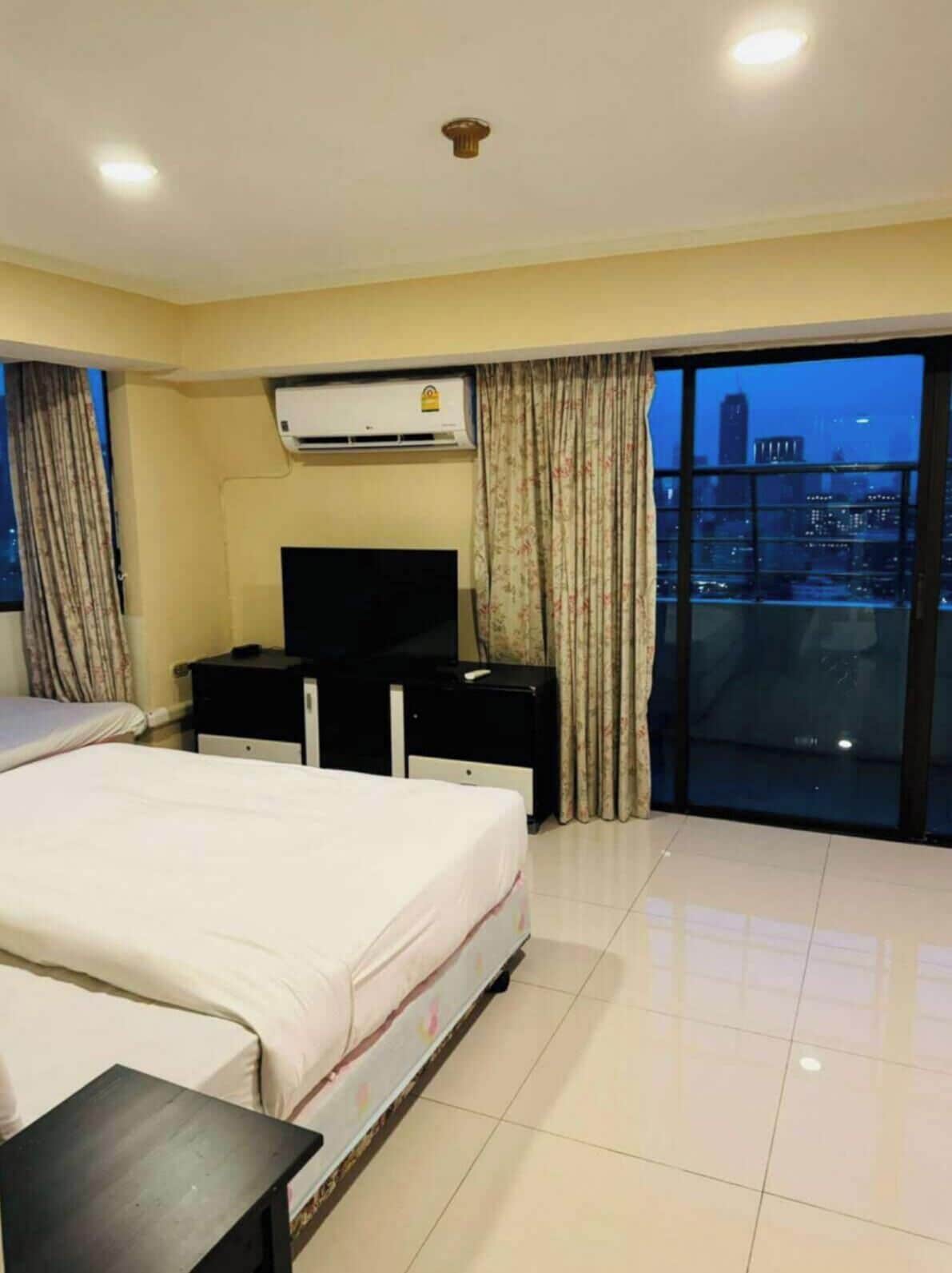 Quarto de hotel com cama, televisão, secretária, unidade de ar condicionado e vista para a paisagem urbana através de uma janela grande.