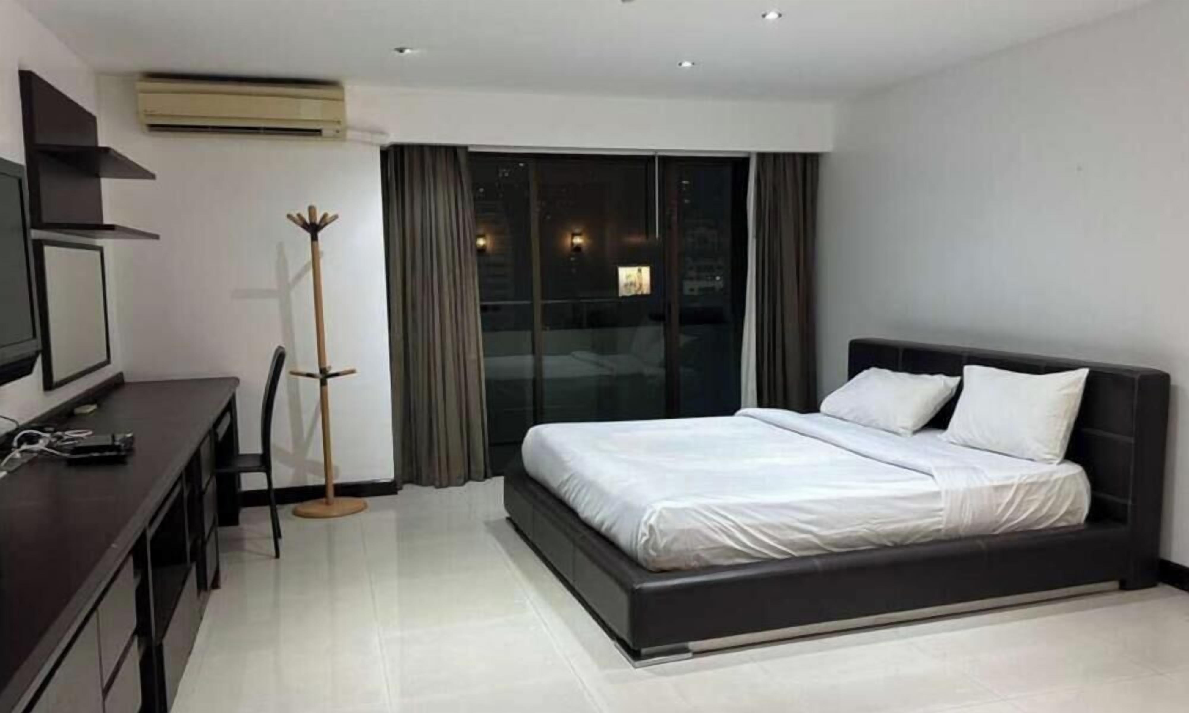 Quarto de hotel moderno com uma cama, escrivaninha, cabideiro e vista da cidade através de amplas janelas.