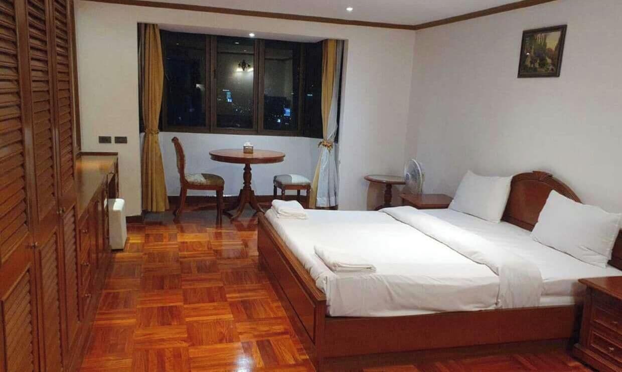 Quarto de hotel com piso de madeira, uma cama, uma mesa de jantar com cadeiras, uma mesinha com um ventilador e uma janela com cortinas.