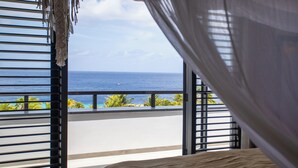 Uma varanda com vista para o mar, uma cama com colcha branca e uma cortina entreaberta.