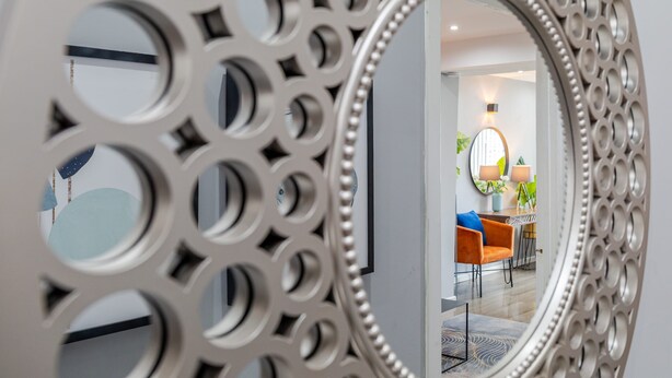 Um espelho decorativo com padrão geométrico, refletindo um quarto com design de interiores moderno, incluindo um sofá e um abajur.