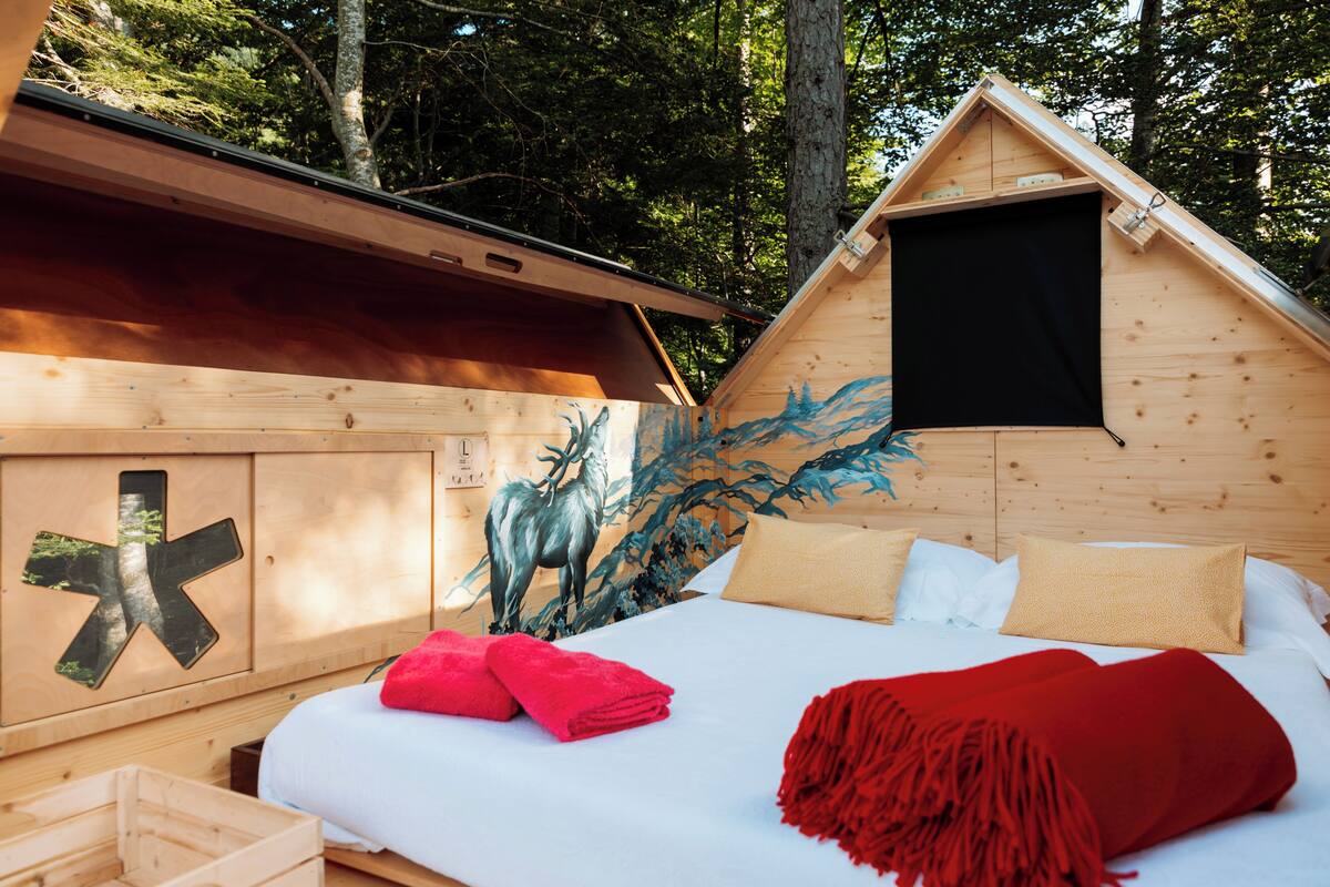 Uma cabana de madeira com uma cama, roupa de cama vermelha e branca, e um mural de um veado.