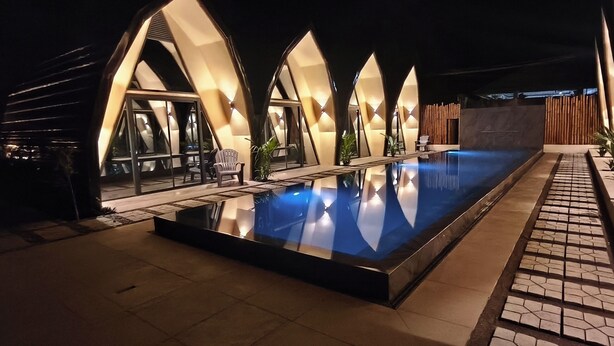 Piscina com deck revestido, cercada por arquitetura moderna com estruturas arqueadas e iluminação ambiente.