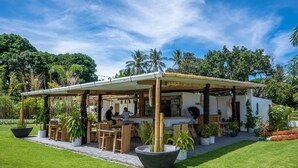 Uma área de estar coberta ao ar livre, com mesas e cadeiras, cercada por plantas em vasos e um edifício branco.