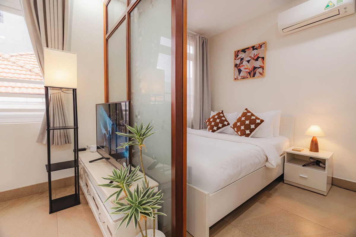 Quarto de hotel com cama, mesa de cabeceira, luminária, TV e plantas em vasos.