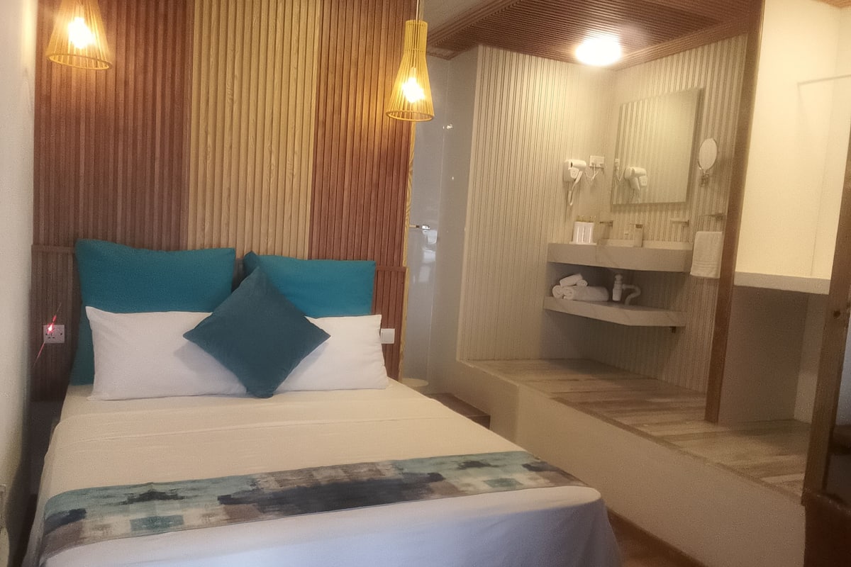 Cama bem arrumada com almofadas, mesa de cabeceira, luminária pendente e banheiro com espelho e porta-toalhas.