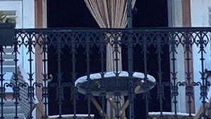 Uma varanda com mesa e cadeiras, um guarda-sol branco e uma janela com cortinas.