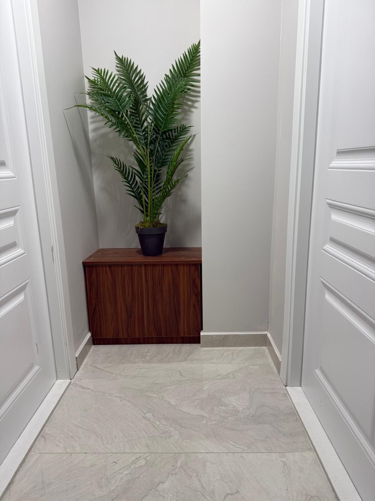 Um corredor estreito com uma planta em vaso sobre um banco de madeira, paredes brancas e chão de mármore.
