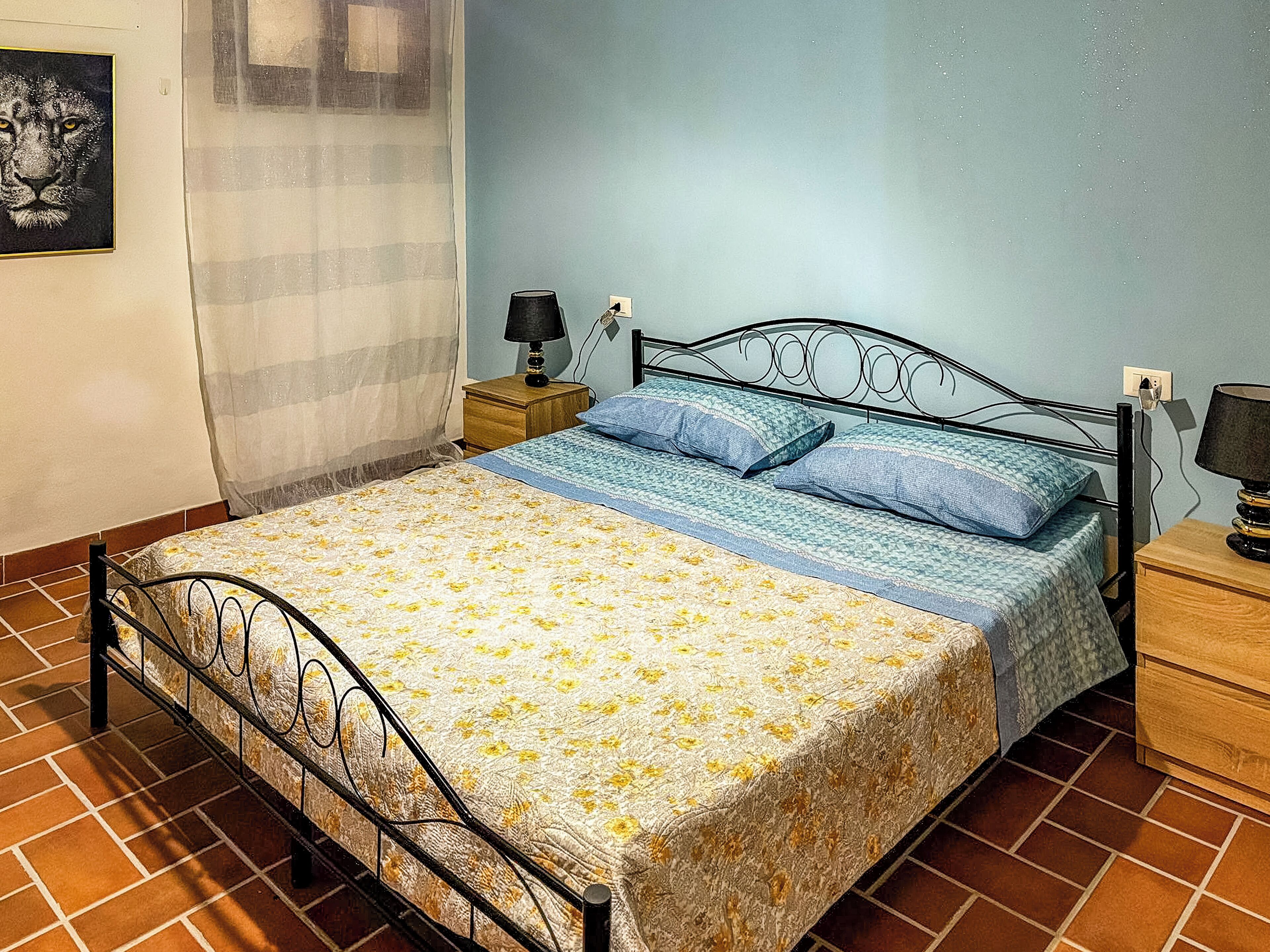 Um quarto com estrutura de cama de metal, colcha floral, duas almofadas, mesa de cabeceira com abajur e um quadro na parede.