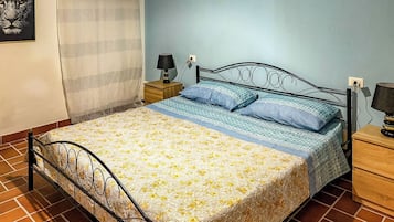 Um quarto com estrutura de cama de metal, colcha floral, duas almofadas, mesa de cabeceira com abajur e um quadro na parede.