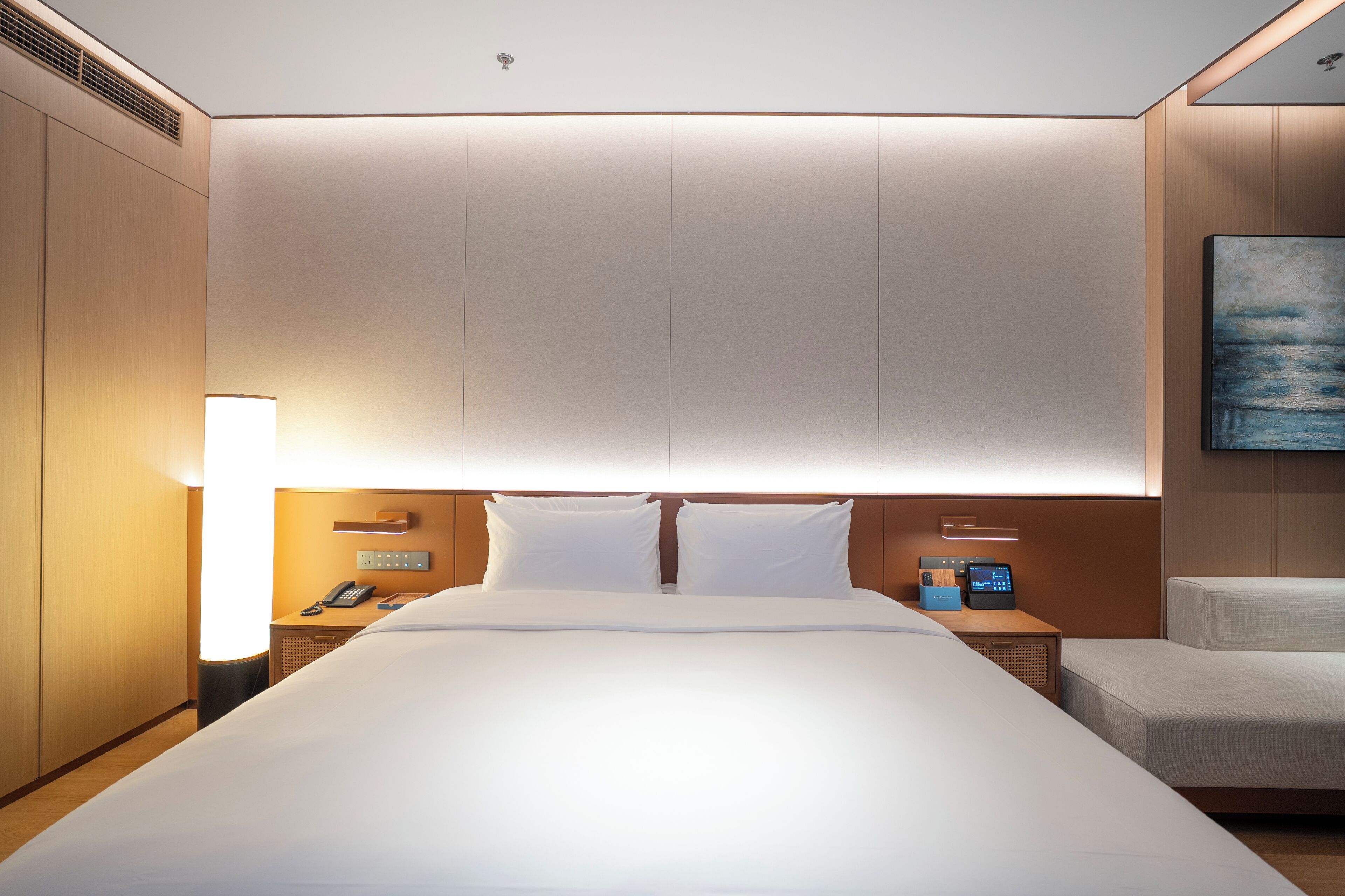 Quarto de hotel com uma cama grande, mesas de cabeceira, uma luminária, um relógio digital e um quadro na parede.
