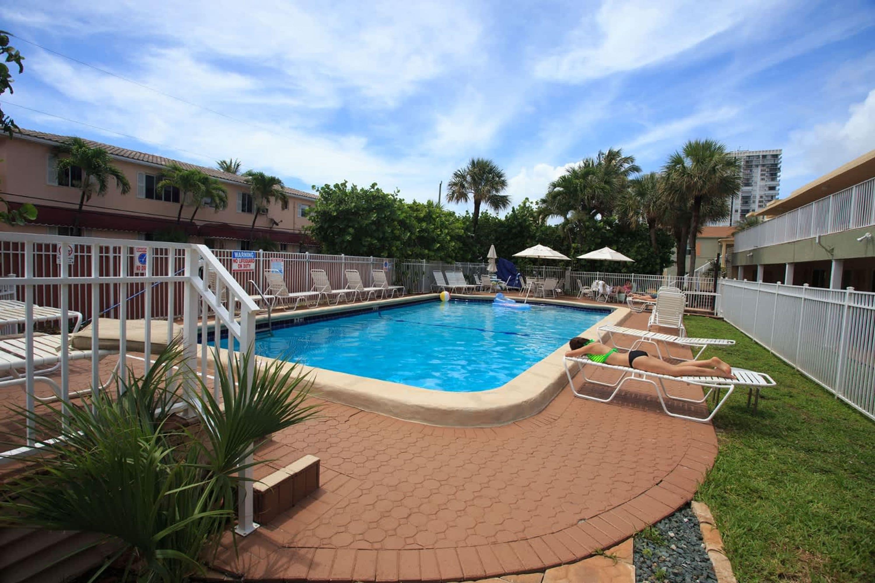 Piscina com espreguiçadeiras, área cercada e um edifício ao fundo.