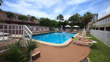Piscina com espreguiçadeiras, área cercada e um edifício ao fundo.