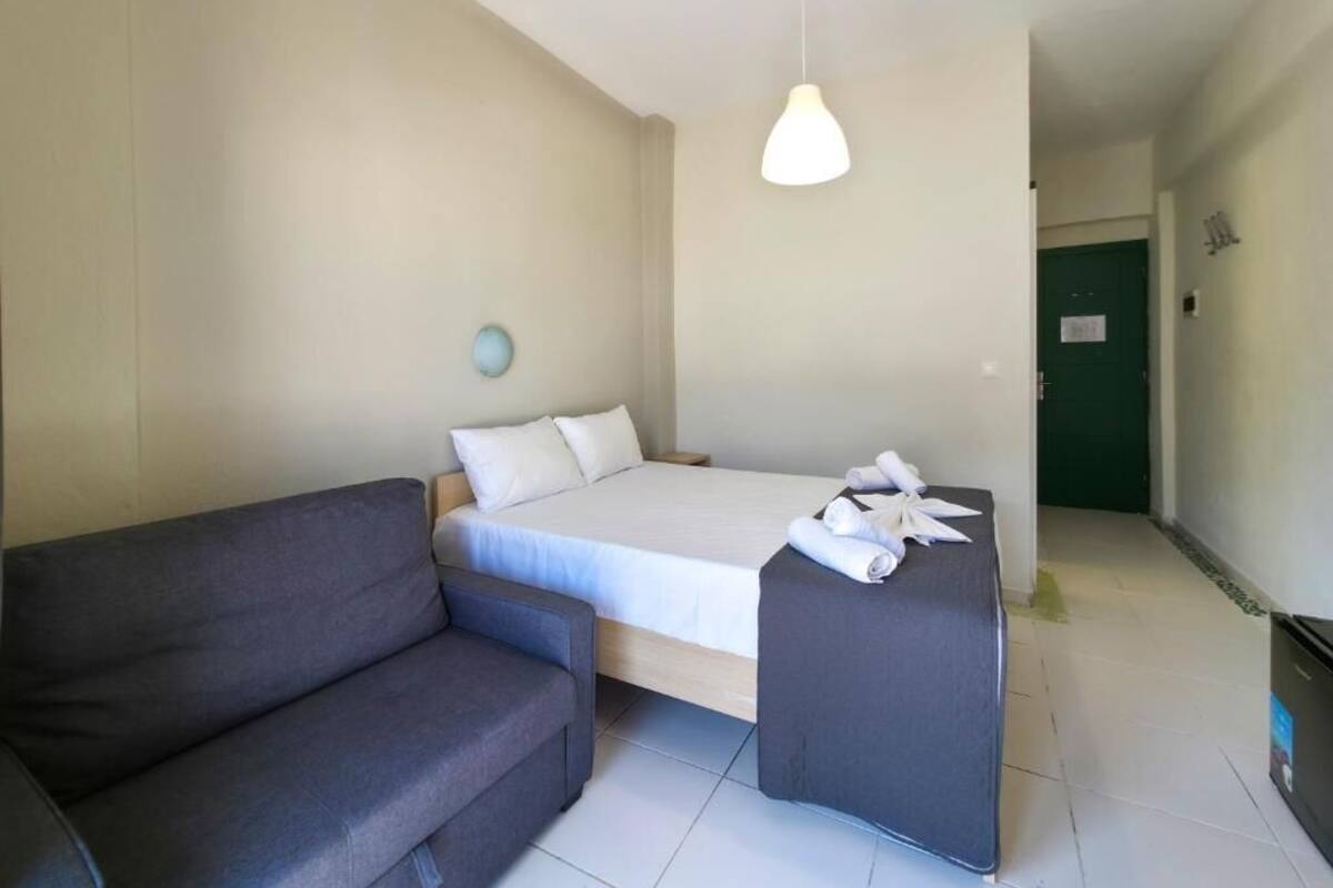 Quarto com cama, sofá, pequena mesa, luminária pendente e uma porta ao fundo.