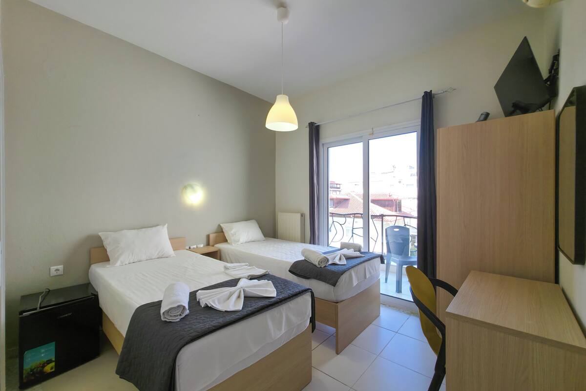 Quarto de hotel com duas camas, uma escrivaninha, uma cadeira, televisão e varanda com vista.