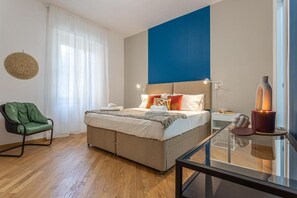Quarto com cama, cadeira, mesa de cabeceira, abajur e janela com cortinas.