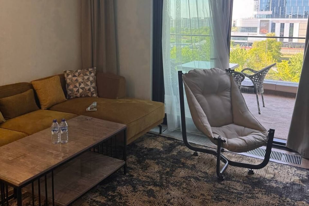 Quarto de hotel com sofá, uma cadeira, uma mesinha e vista para a cidade através de grandes janelas.