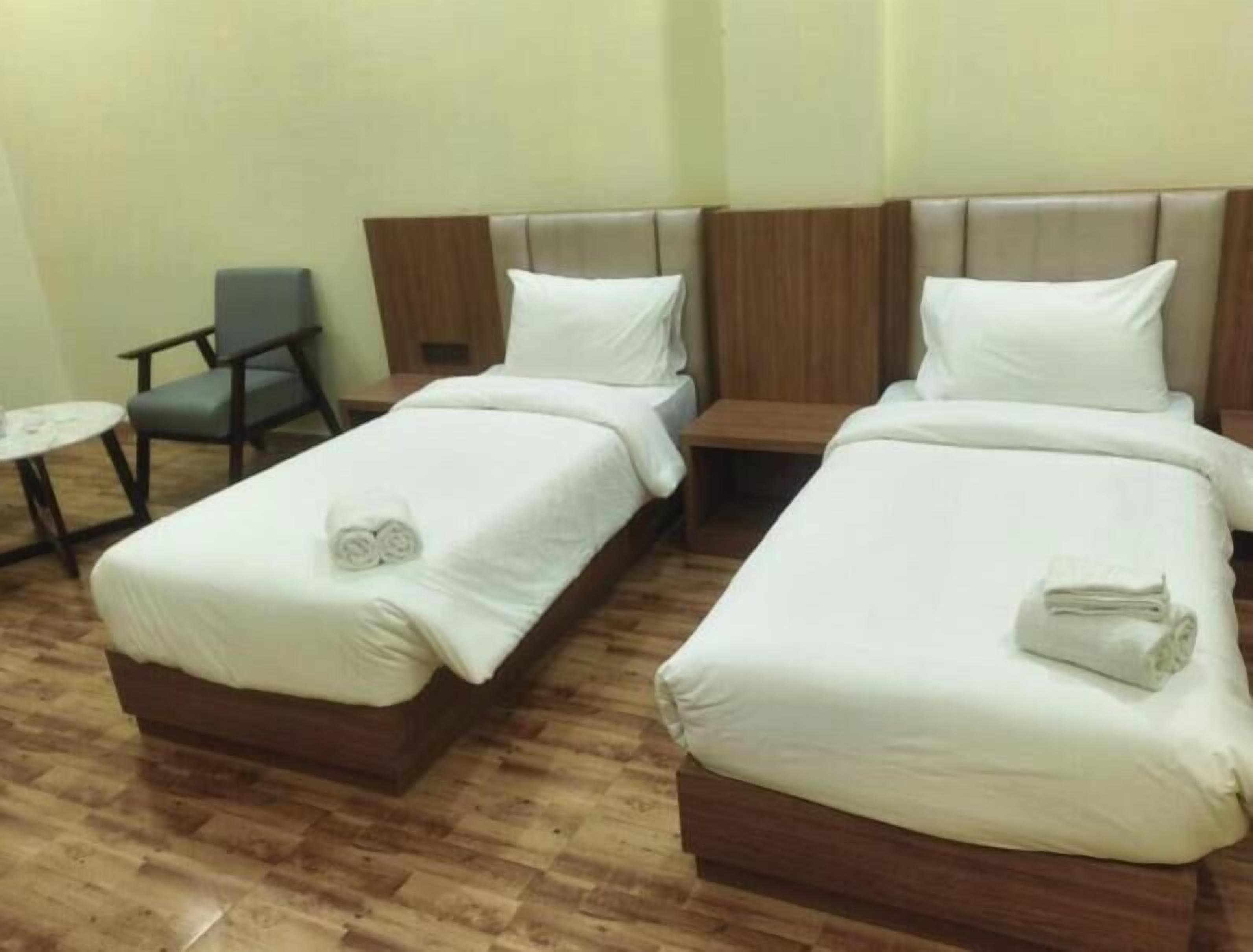 Quarto de hotel com duas camas de solteiro, uma mesa pequena, uma cadeira e piso de madeira.