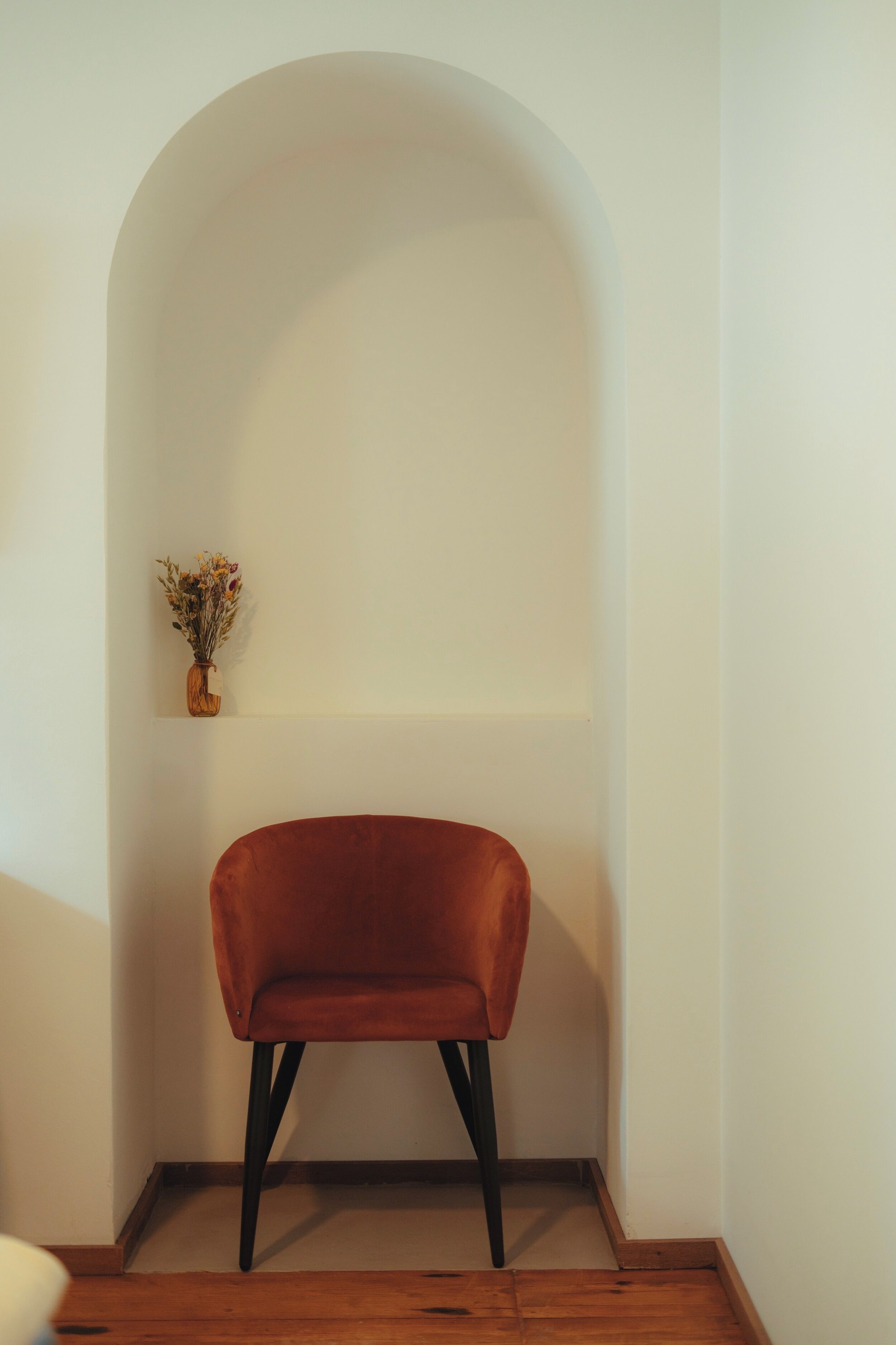 Quarto de canto com uma cadeira vermelha, piso de madeira e um pequeno vaso com flores secas na parede.