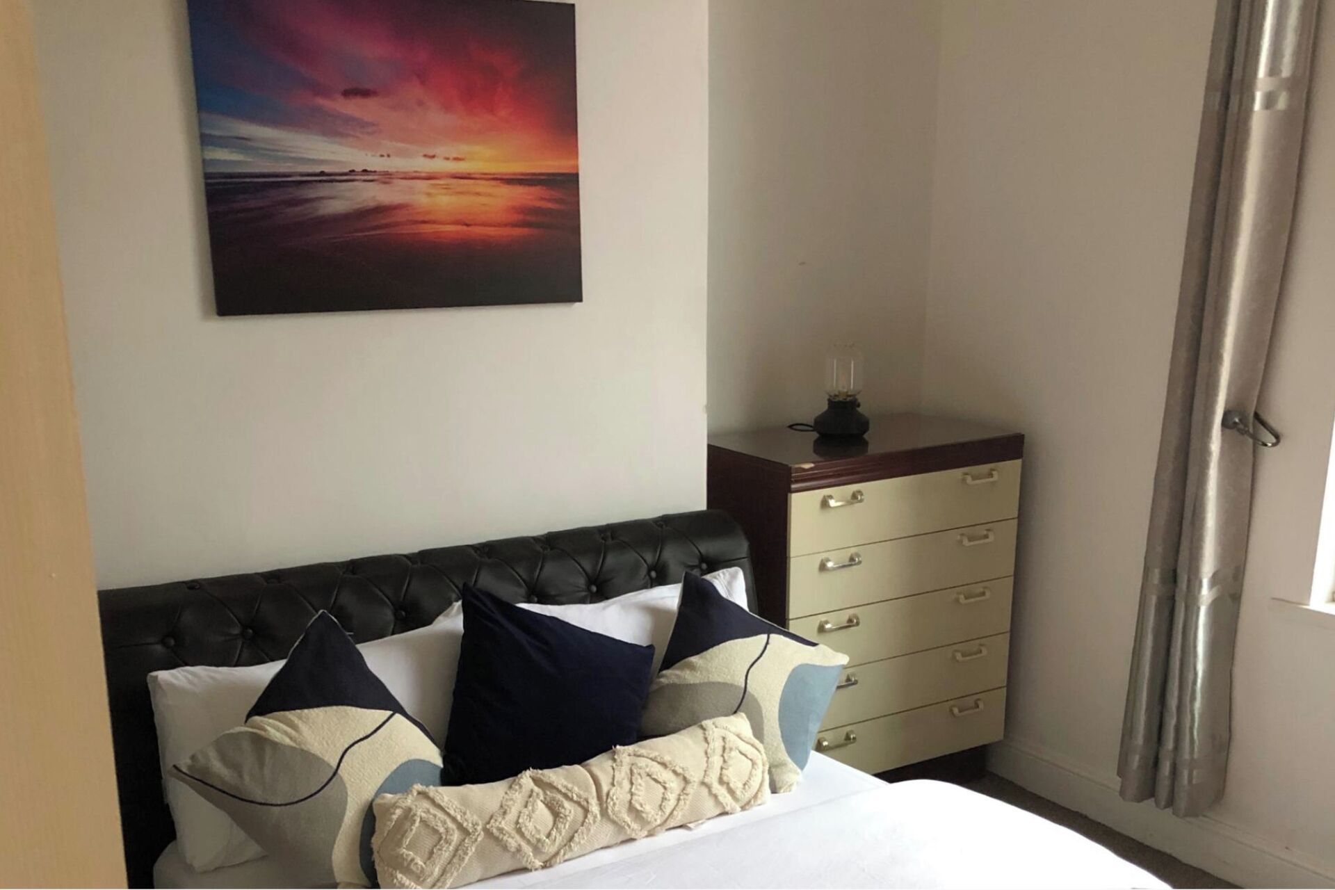 Quarto com cama, mesa de cabeceira, cômoda e pintura de um pôr do sol.