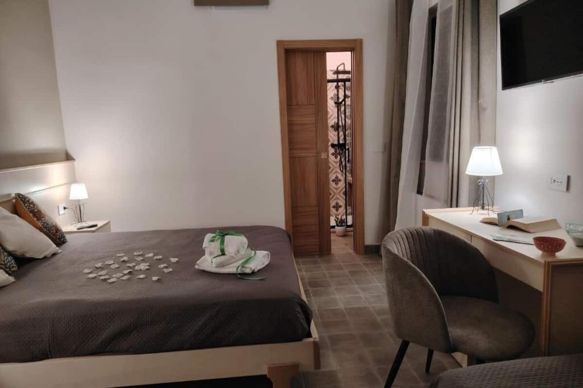 Quarto de hotel com cama, escrivaninha, cadeira, televisão e porta para uma varanda.