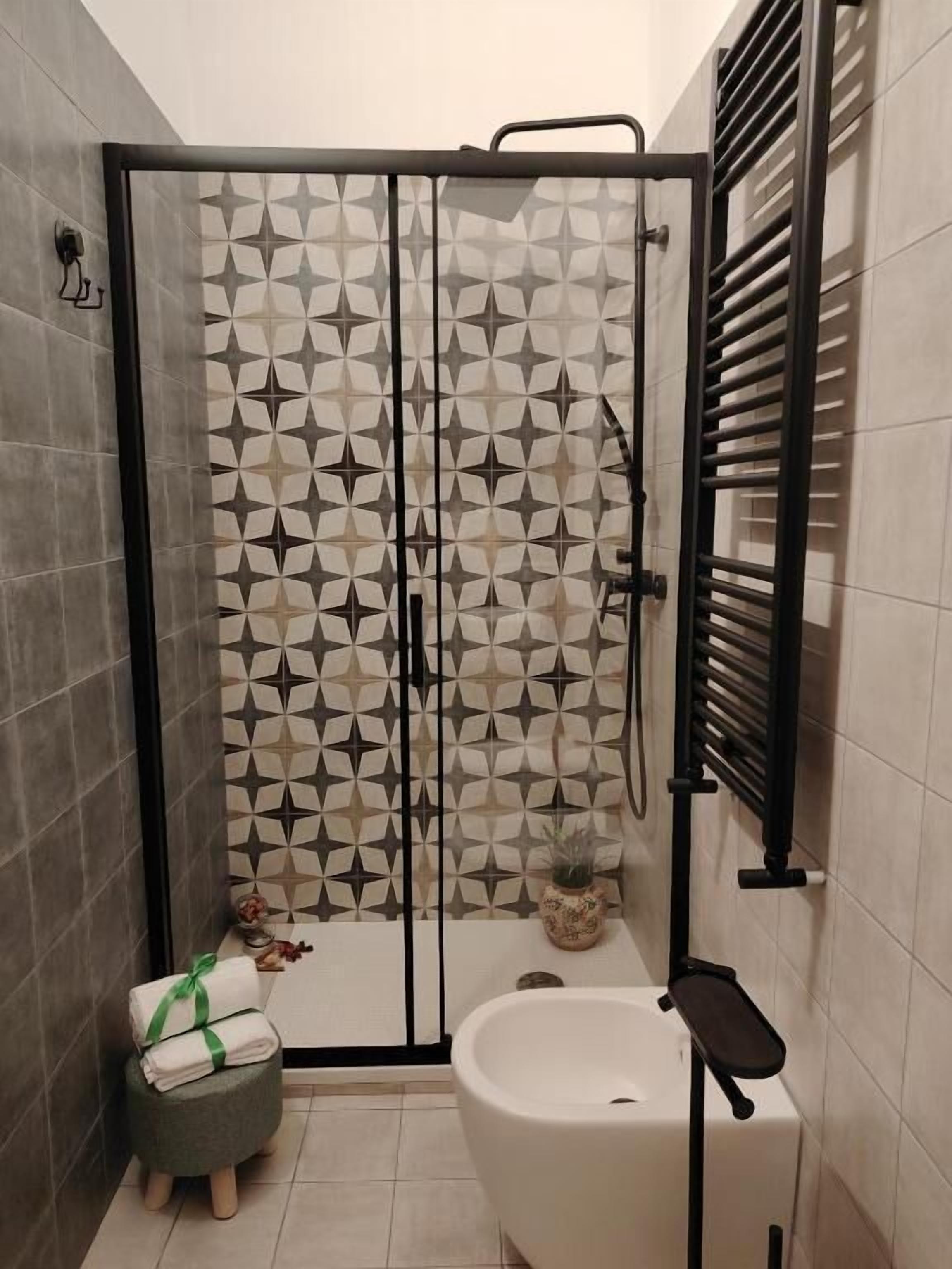 Um chuveiro com padrão geométrico de azulejos, uma banheira branca e uma toalha sobre um banquinho.