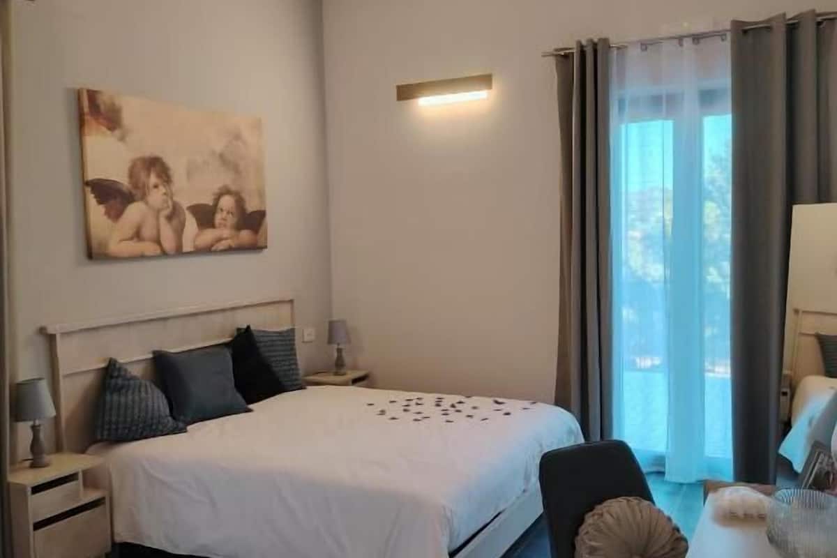 Quarto de hotel com cama, mesas de cabeceira, escrivaninha com cadeira, um quadro na parede e janela com cortinas.