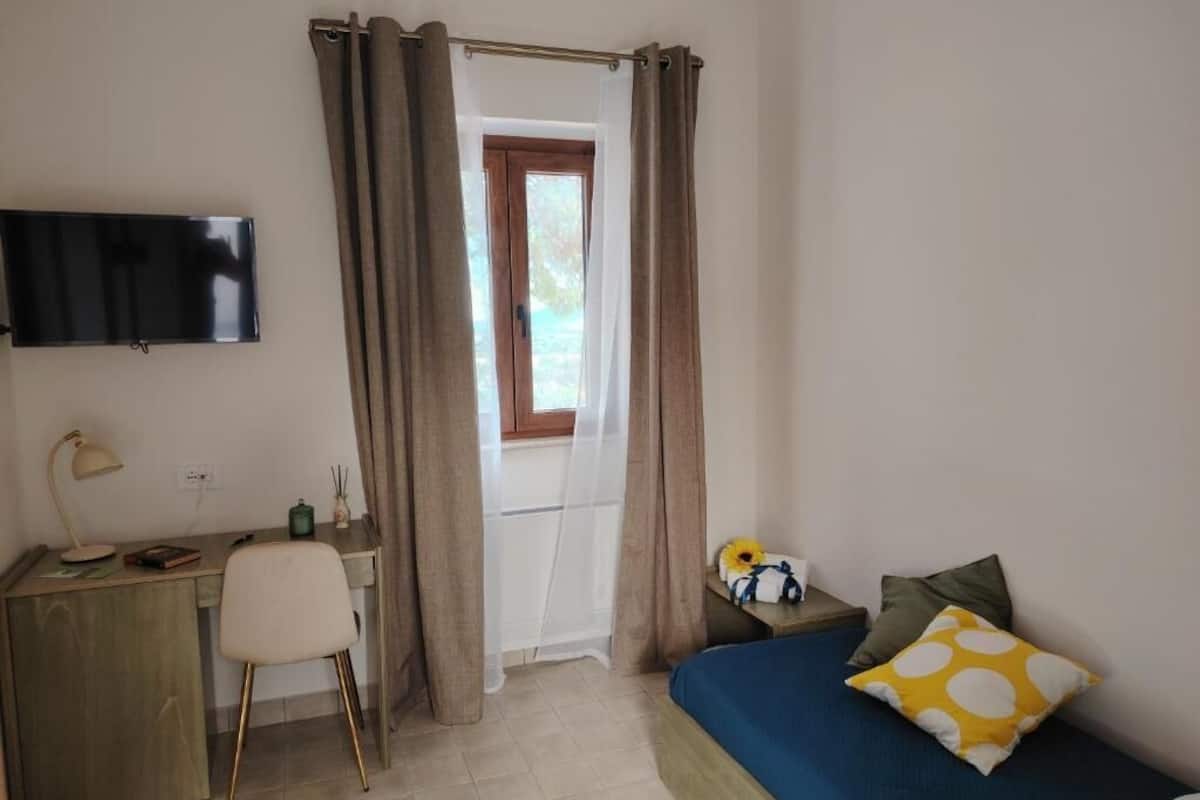 Quarto com cama, escrivaninha, cadeira, televisão e janela com cortinas.