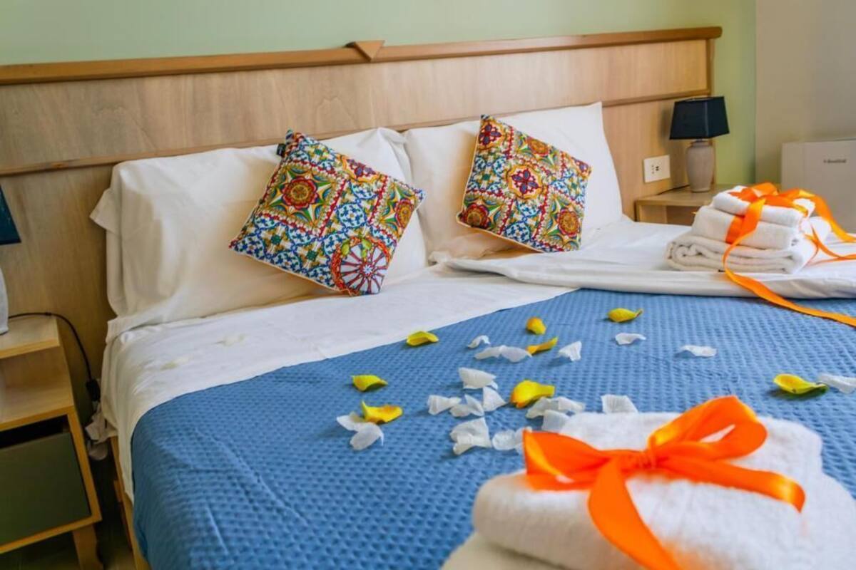 Quarto de hotel com cabeceira de madeira, colcha azul com textura, travesseiros brancos e cama bem arrumada com toalhas e uma cesta de frutas.