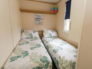Um quarto pequeno e acolhedor com duas camas, uma prateleira na parede com malas coloridas, uma janela com cortina e uma placa na parede.