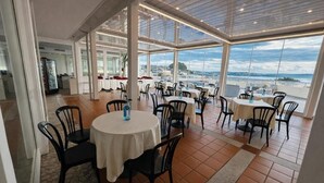 Um restaurante com mesas e cadeiras redondas, grandes janelas e vista para o mar.