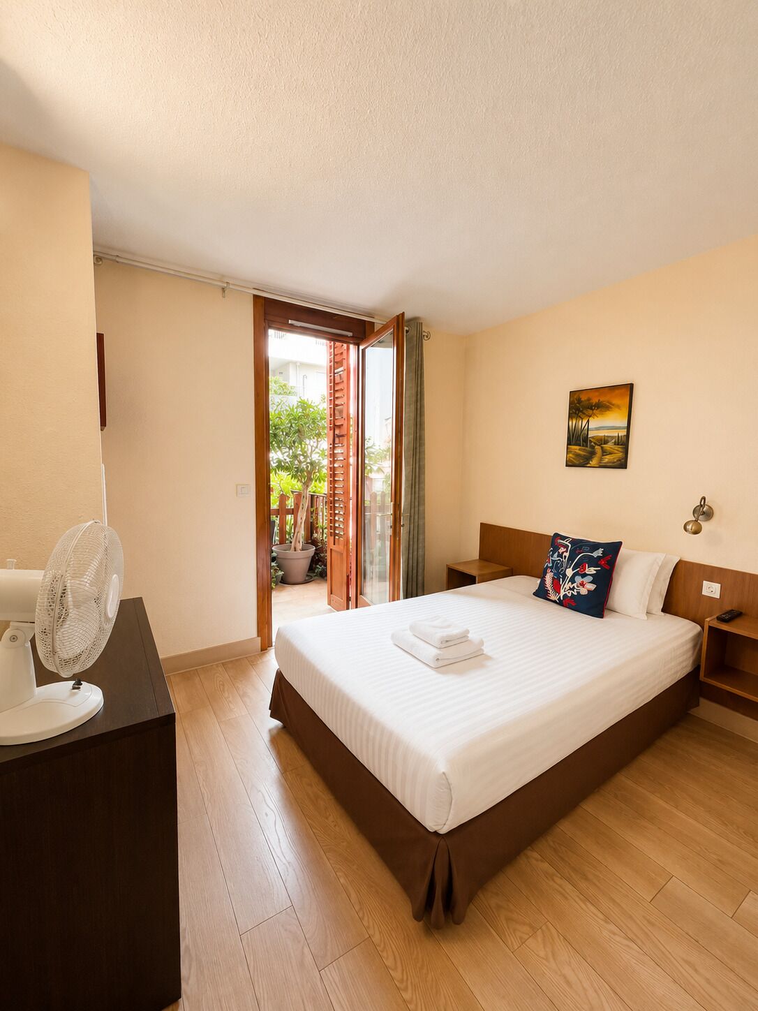 Quarto de hotel com cama, ventilador, mesa de cabeceira, pintura e vista para o exterior.