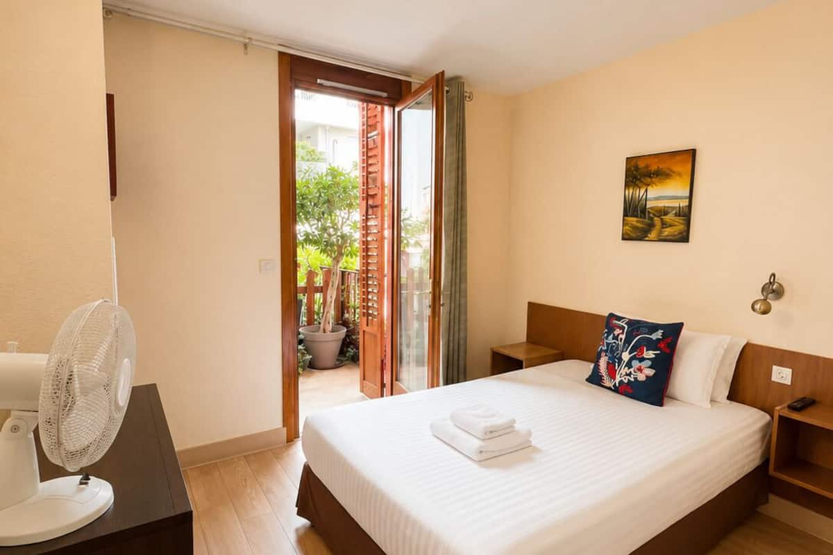 Quarto de hotel com cama, ventilador, mesa de cabeceira, pintura e vista para o exterior.