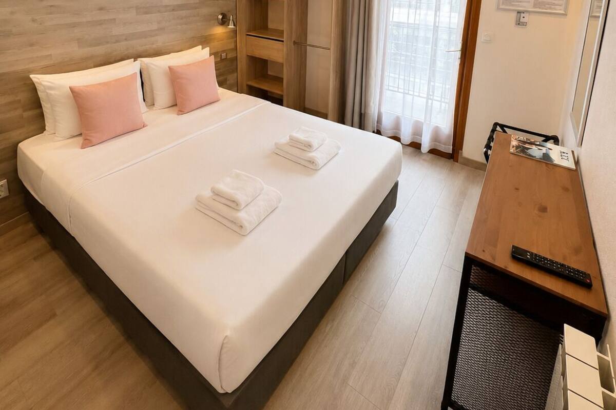Quarto de hotel com cama, guarda-roupa de madeira, escrivaninha com controle remoto e janela com cortinas.