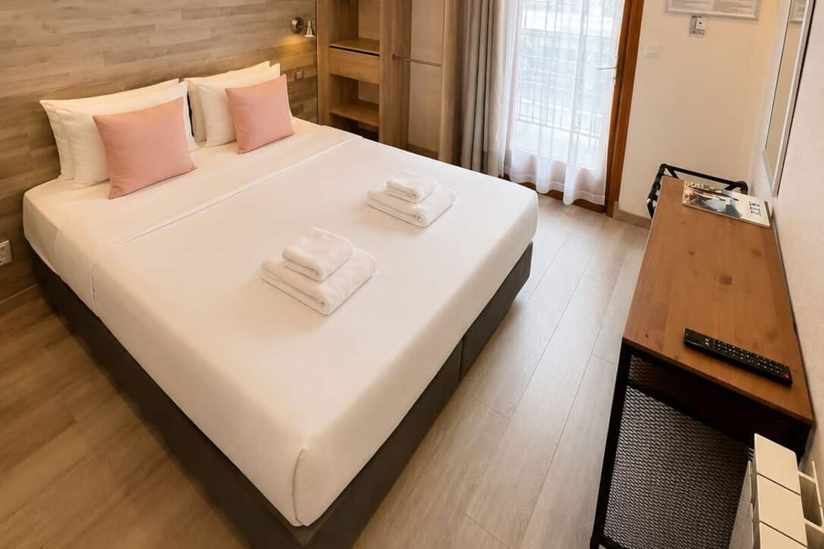 Quarto de hotel com cama, guarda-roupa de madeira, escrivaninha com controle remoto e janela com cortinas.