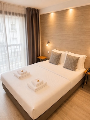 Quarto de hotel com uma cama grande, mesa de cabeceira, luz acoplada à parede e vista através de cortinas finas.